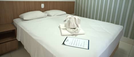 Uma cama bem arrumada com lençóis brancos, uma toalha dobrada e um travesseiro. Há uma mesa de cabeceira de madeira ao lado da cama, e uma televisão de tela plana fixada na parede acima.