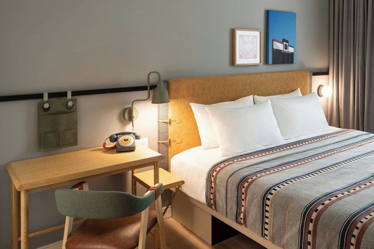 Quarto de hotel com uma cama, escrivaninha, cadeira, luminária e prateleira na parede.