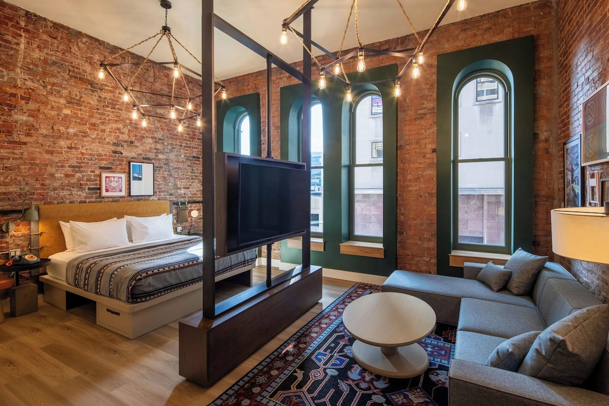 Quarto de hotel moderno com paredes de tijolo aparente, uma cama grande com televisão, um sofá e uma pequena mesa.
