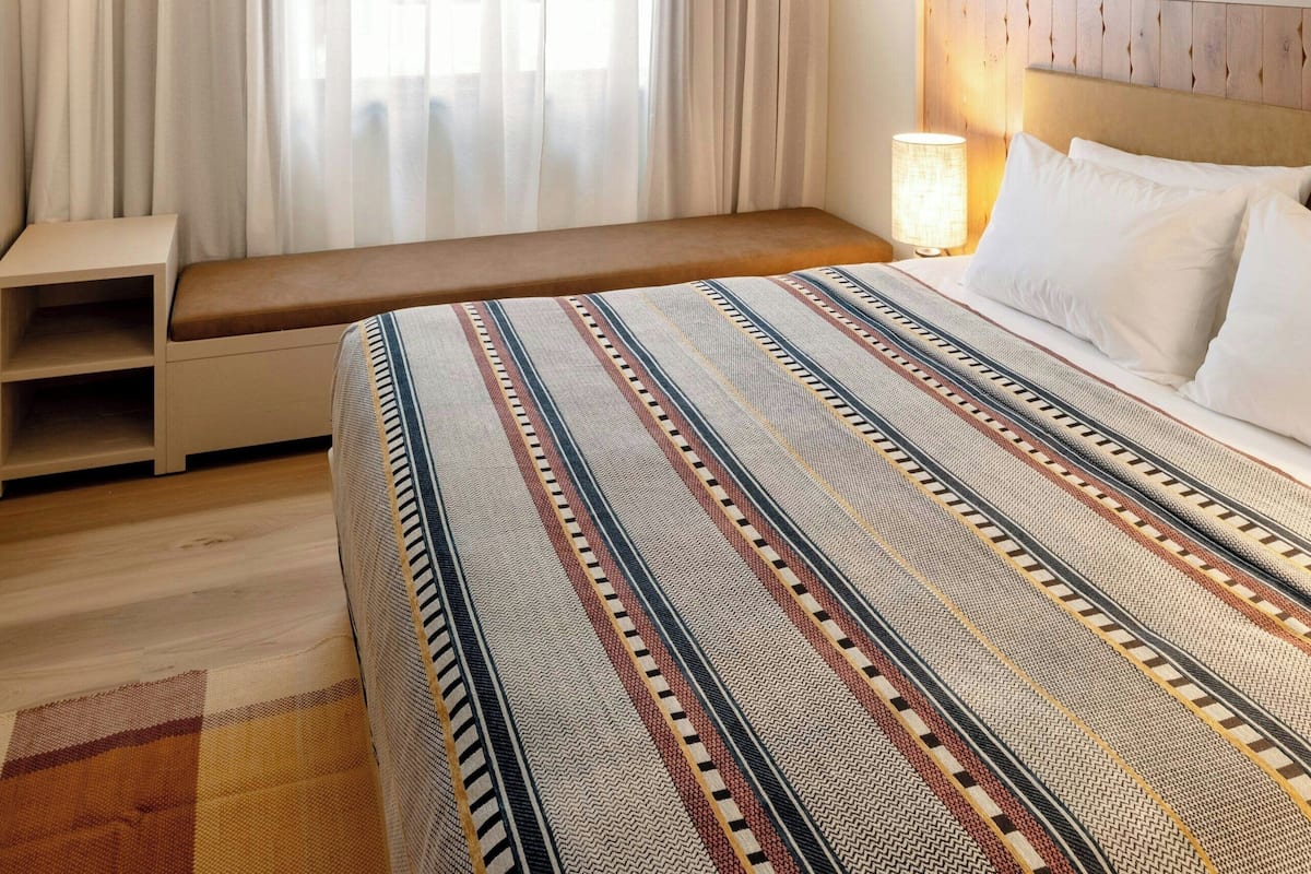 Quarto de hotel com cama, um banco, mesa de cabeceira, luminária e janela com cortinas.