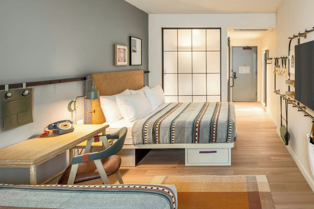 Quarto de hotel moderno com cama, escrivaninha, cadeira e televisão fixada na parede.