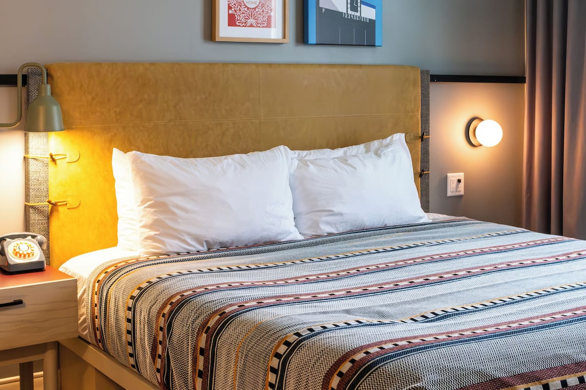Uma cama bem arrumada com um cobreleita estampado, uma luminária de mesa e um quadro na parede.
