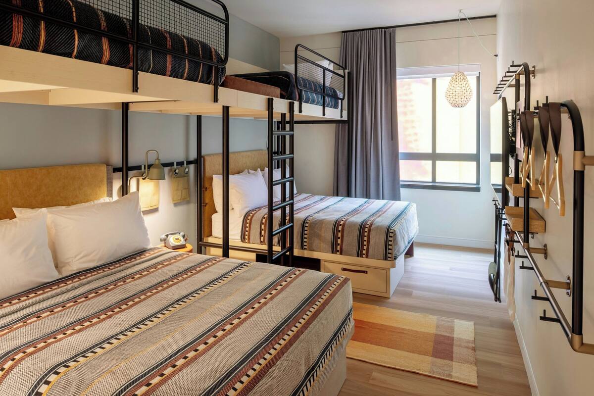 Quarto de hotel com beliches, uma escrivaninha, luminária e janela com cortinas.