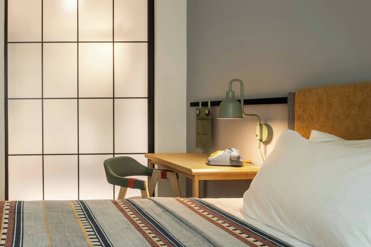 Quarto de hotel com uma cama, uma escrivaninha, uma cadeira, um abajur e uma parede com padrão quadriculado.