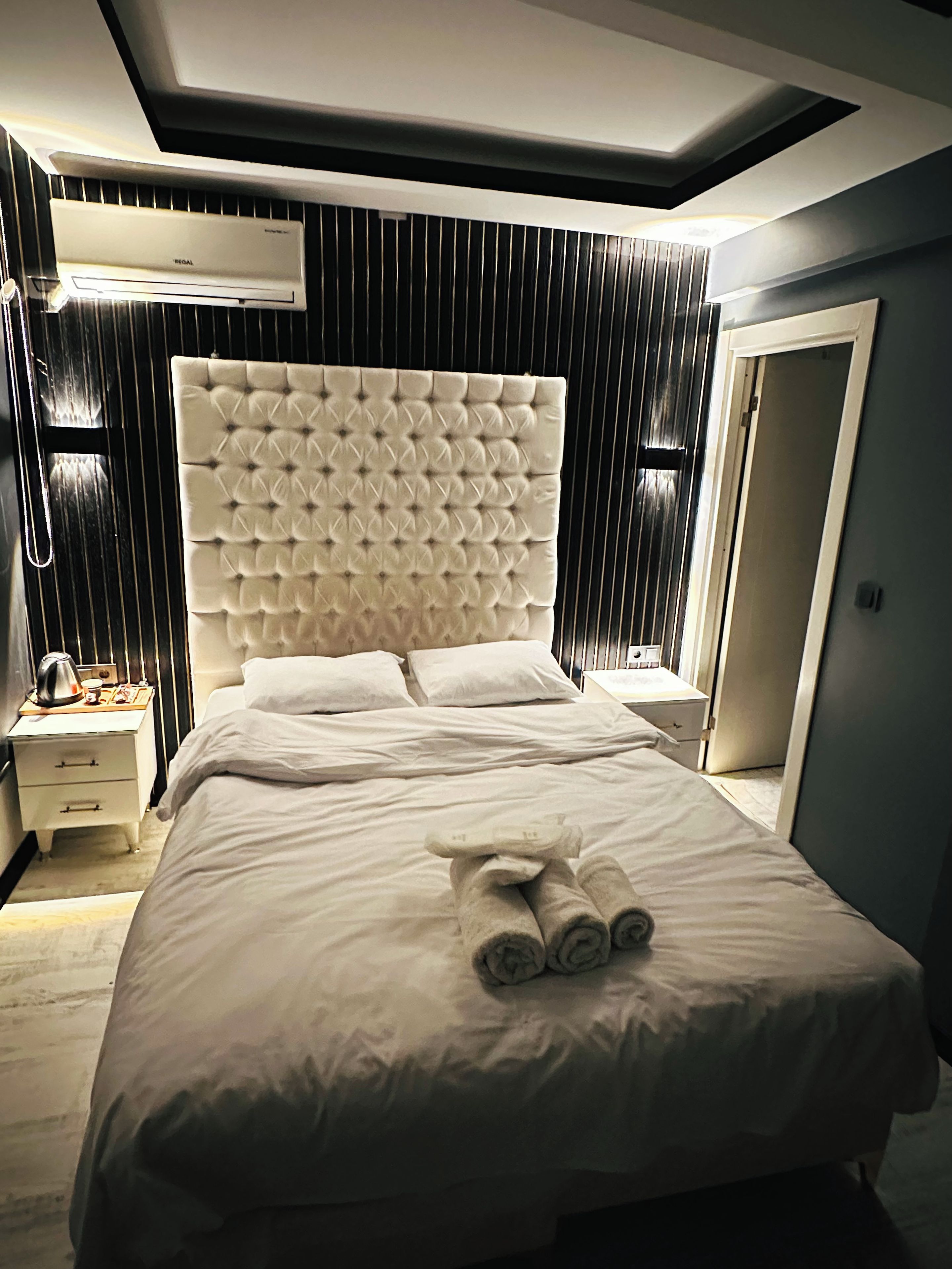 Quarto de hotel com uma cama grande, cabeceira, mesa de cabeceira com abajur e ar condicionado.