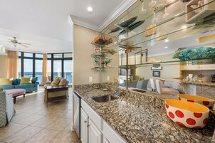 Cozinha moderna com bancadas de granito, pia embutida e prateleira de vidro com objetos decorativos.