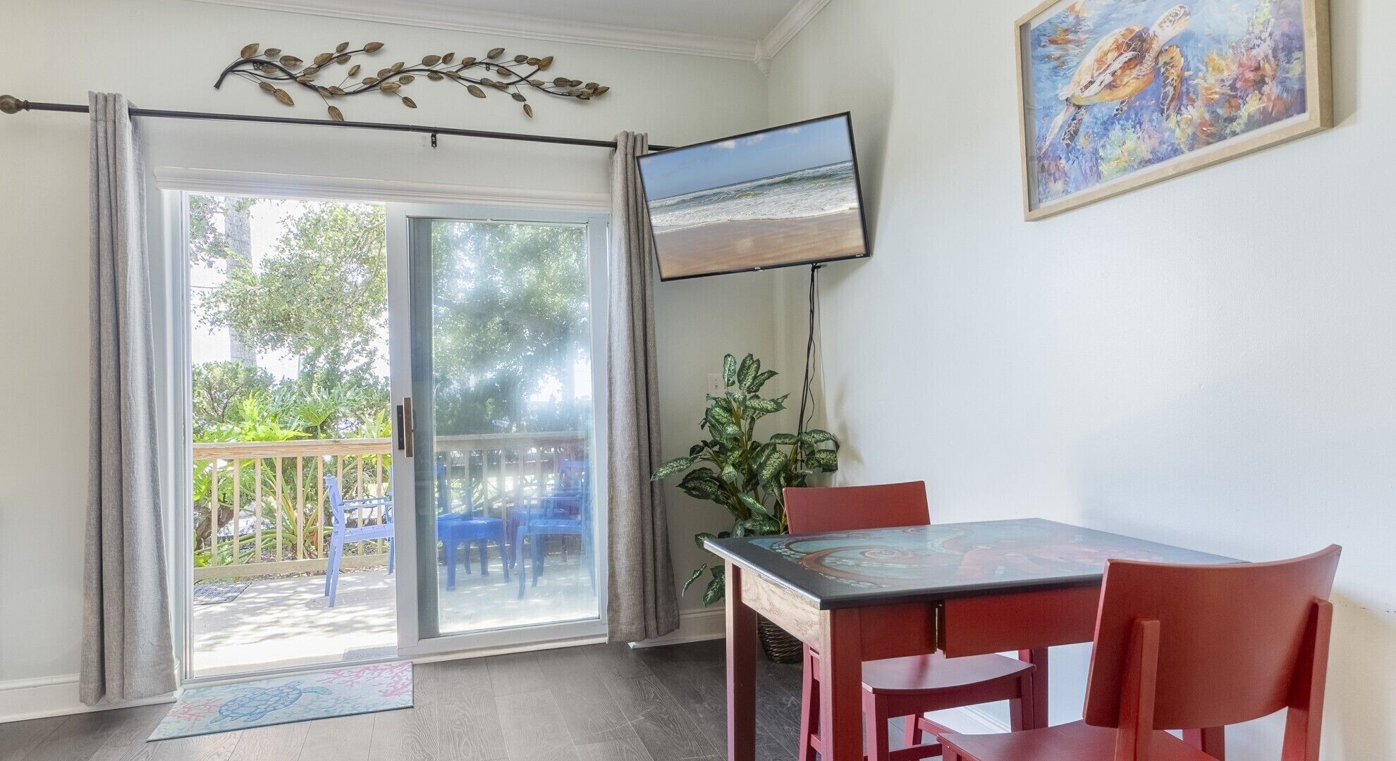 Quarto com porta de vidro deslizante que dá para uma área externa, mesa com cadeiras, planta em vaso e um quadro na parede.