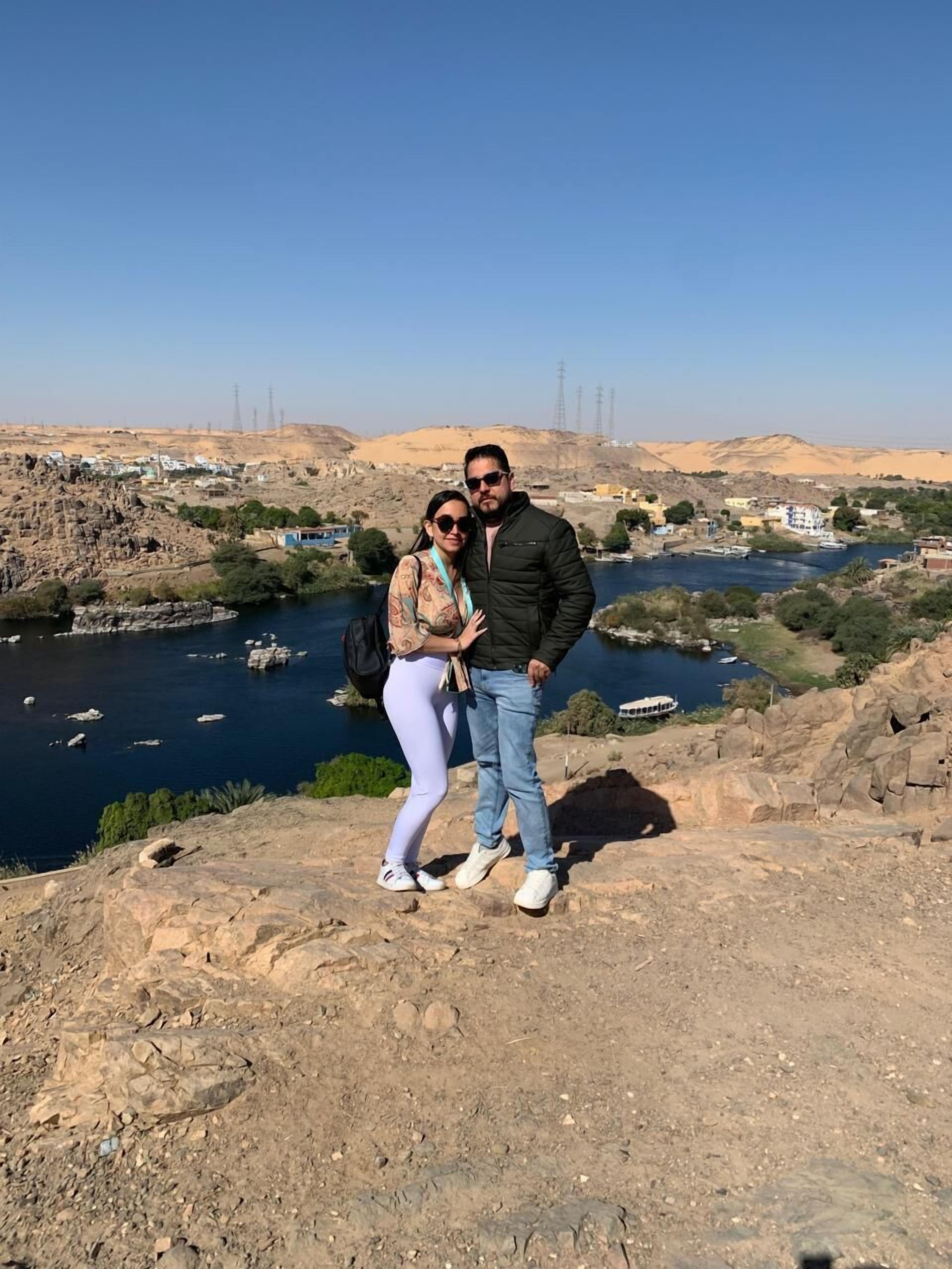 Um casal está em um mirante rochoso, com um rio e edifícios ao fundo.