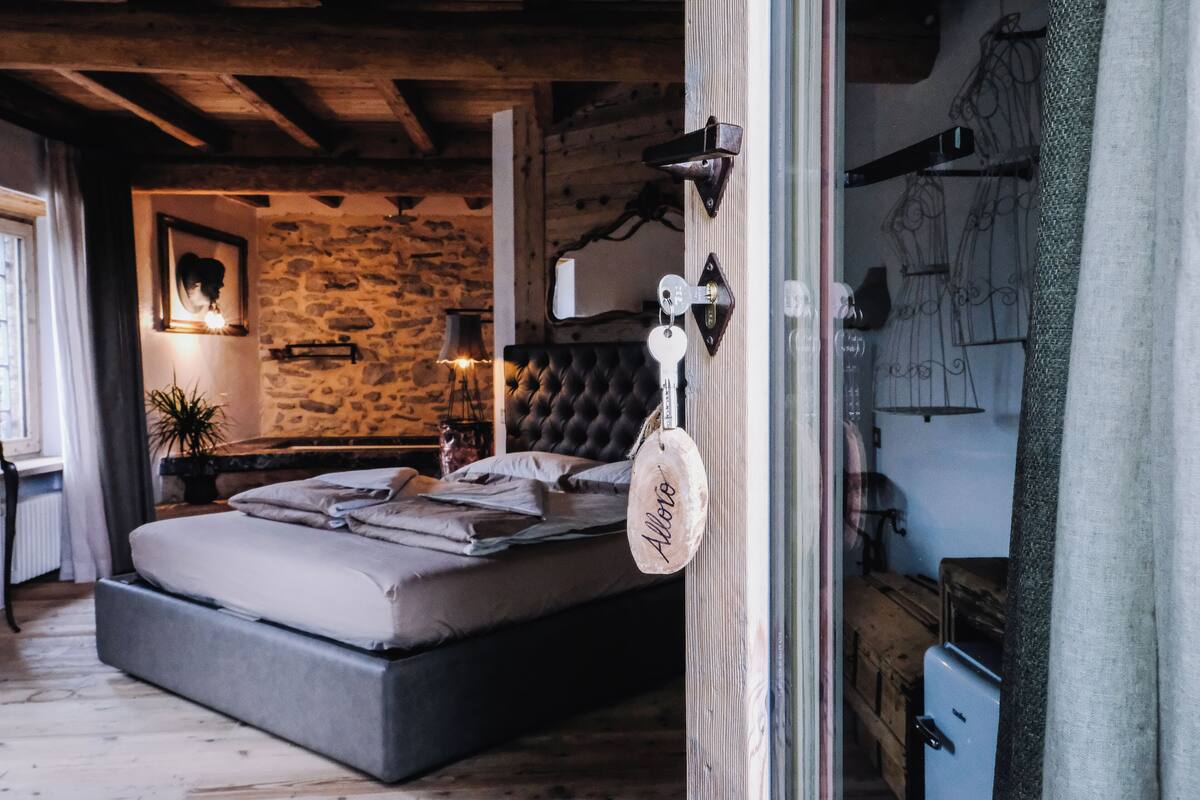 Quarto com parede de pedra, cabeceira estofada, cama com roupa de cama cinza, chave pendurada e cômoda de madeira rústica.