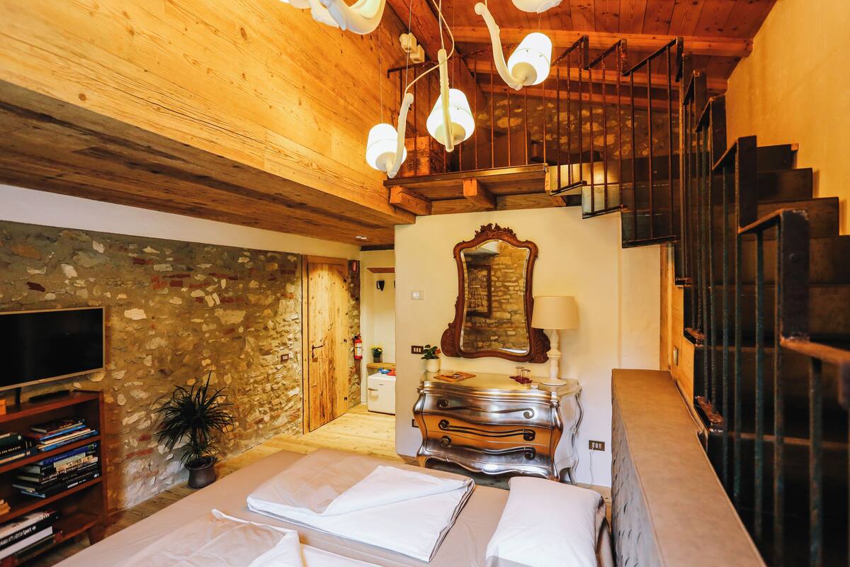 Quarto com teto de madeira, escada, espelho, cômoda e televisão em uma prateleira.