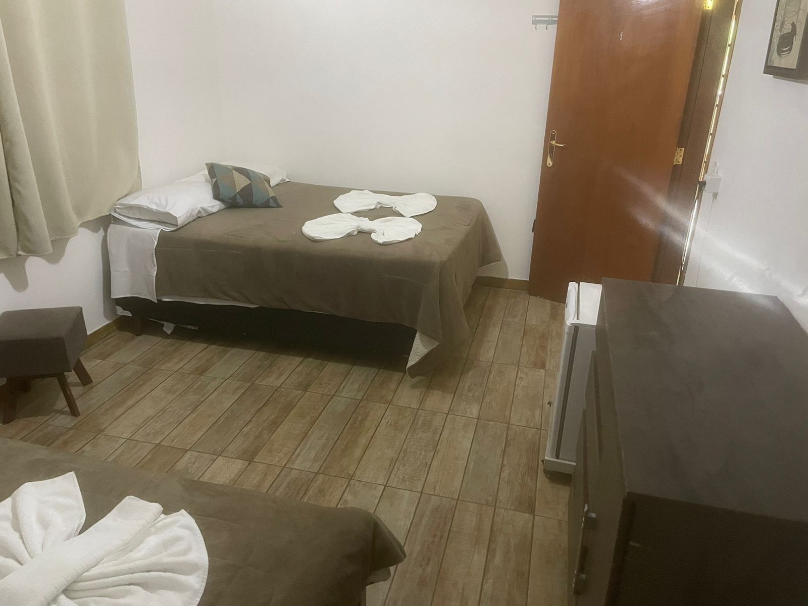 Uma cama de solteiro com colcha marrom e almofadas brancas, uma mesinha de cabeceira pequena e piso de madeira.