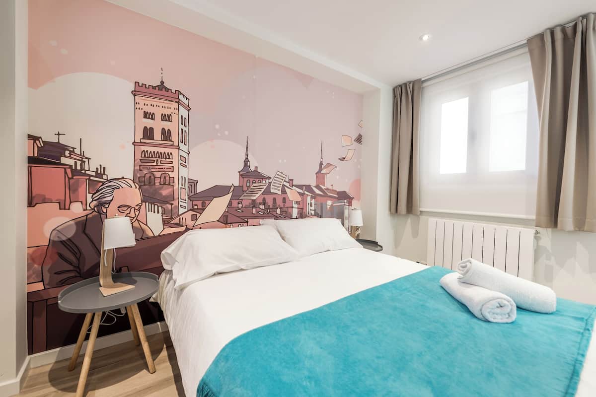 Quarto de hotel com cama, mesa de cabeceira, luminária, janela com cortinas e um mural com vista urbana na parede.