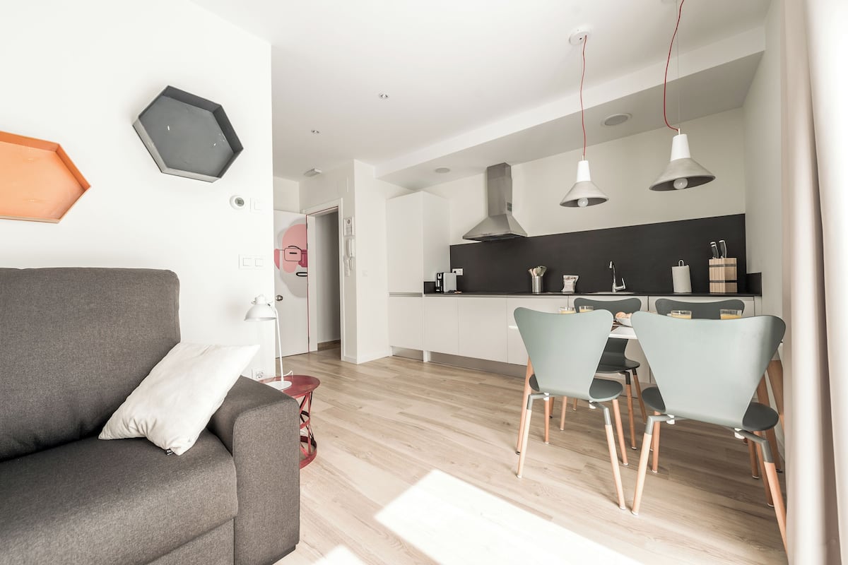 Uma sala de estar moderna com um sofá cinzento, um espaço de refeições com cadeiras azul-claras e uma cozinha ao fundo.