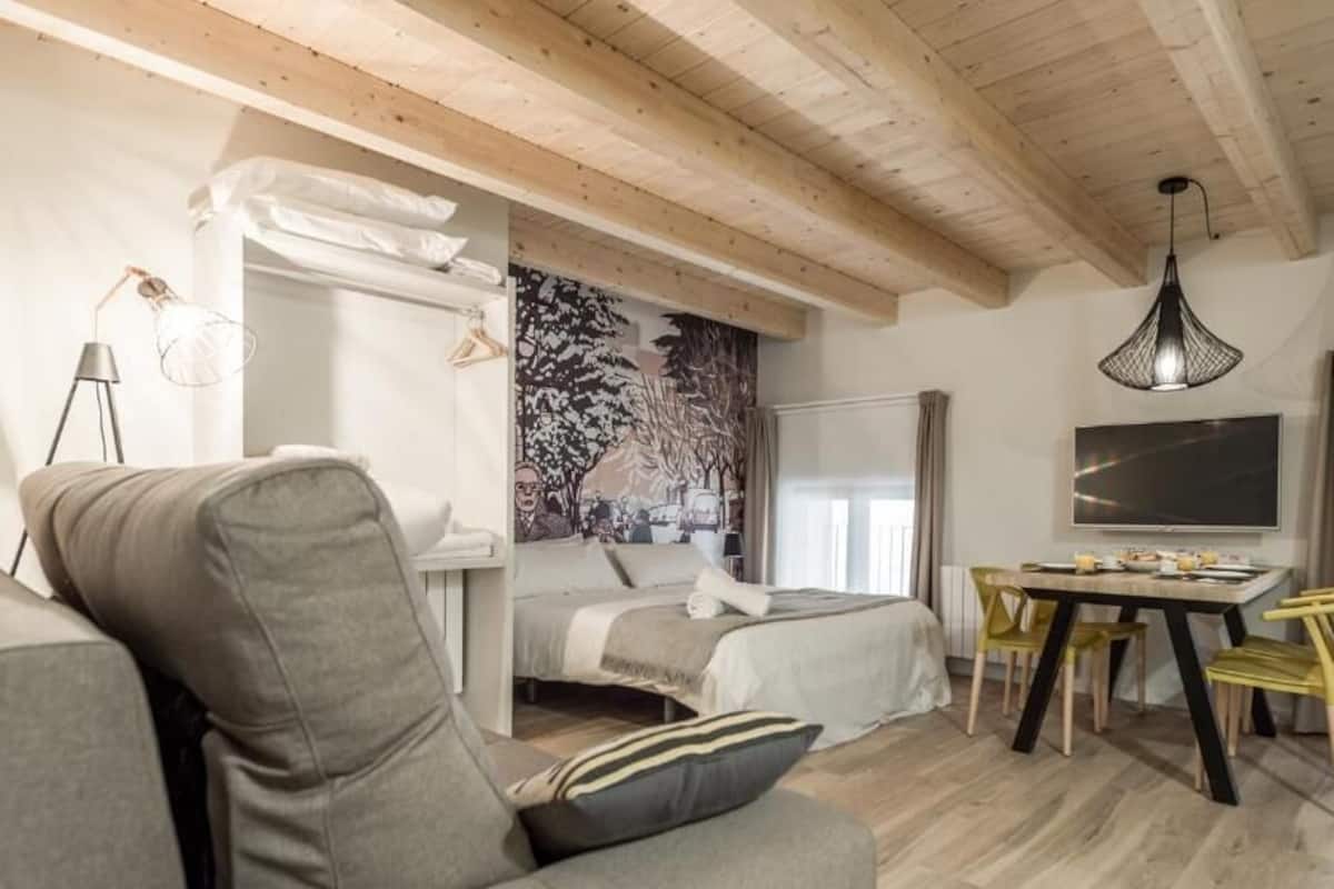 Quarto aconchegante com cama, sofá, mesa de jantar com cadeiras, televisão e uma janela grande.