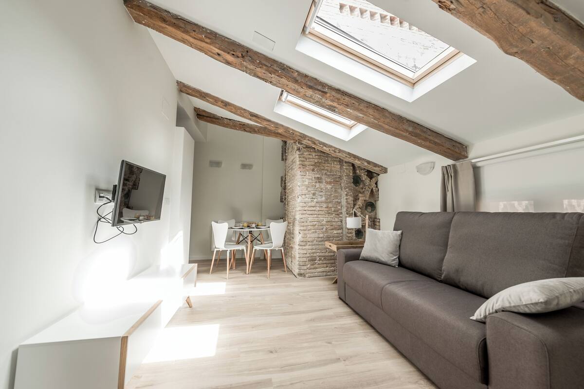 Uma sala de estar moderna com vigas de madeira aparentes, parede de pedra, sofá cinza e área de jantar com mesa e cadeiras brancas.