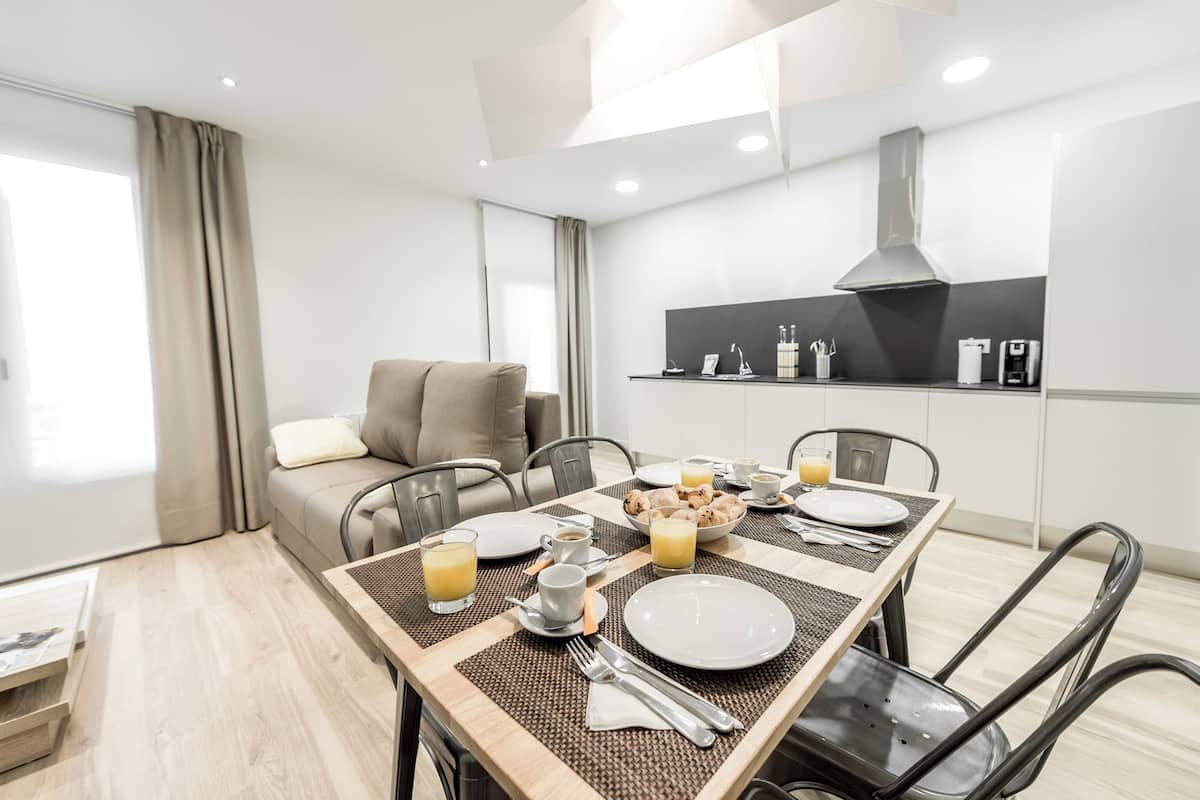 Quarto de hotel moderno com área de refeições, incluindo uma mesa de madeira posta para o café da manhã, um sofá e uma cozinha com bancada preta.