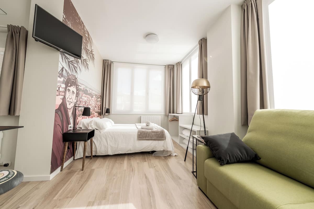 Quarto de hotel moderno com uma cama, um sofá verde, um abajur e uma janela grande com cortinas.