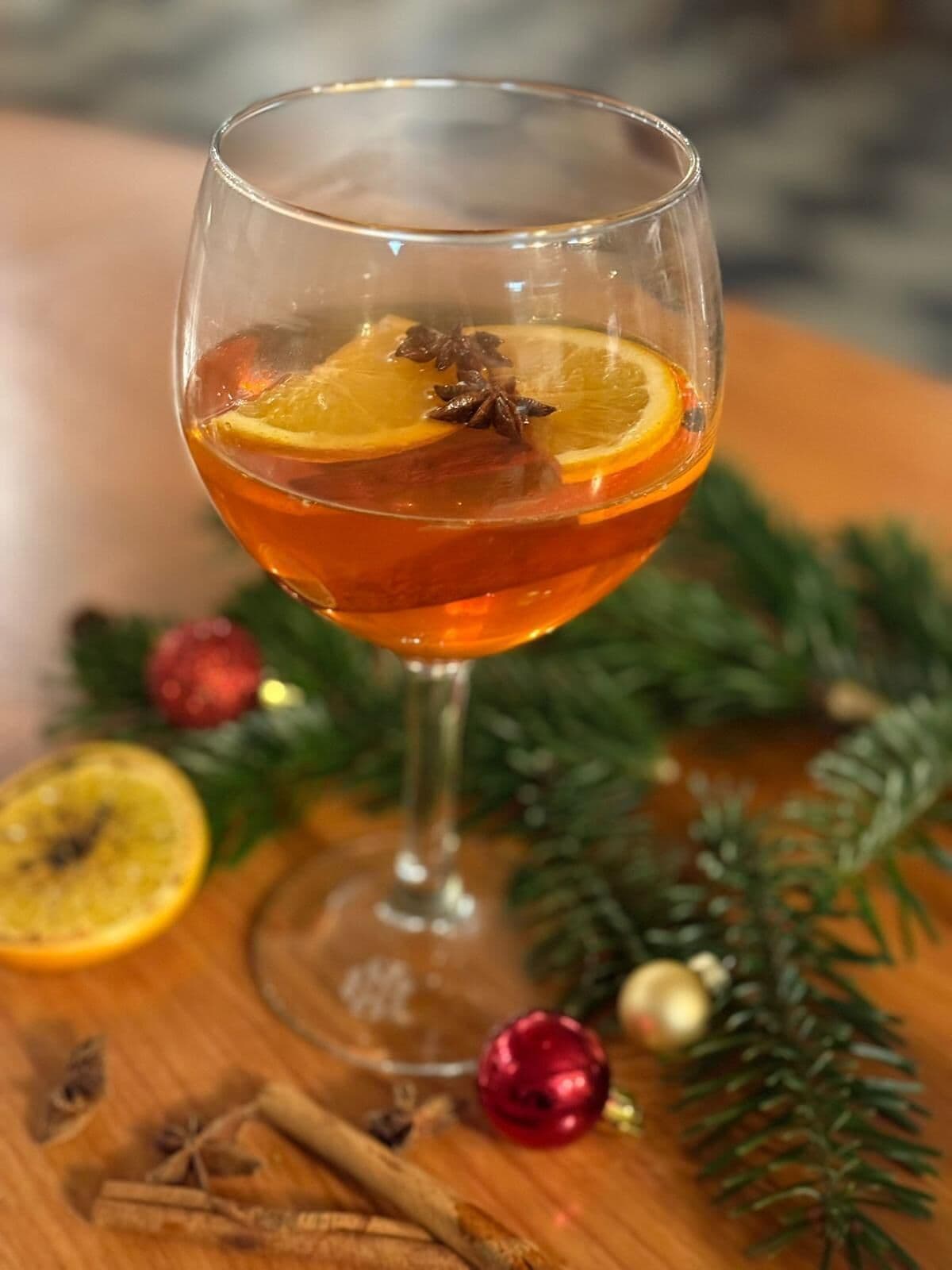Um copo de bebida cor de âmbar decorado com rodelas de laranja e anis estrelado, rodeado de enfeites festivos sobre uma superfície de madeira.