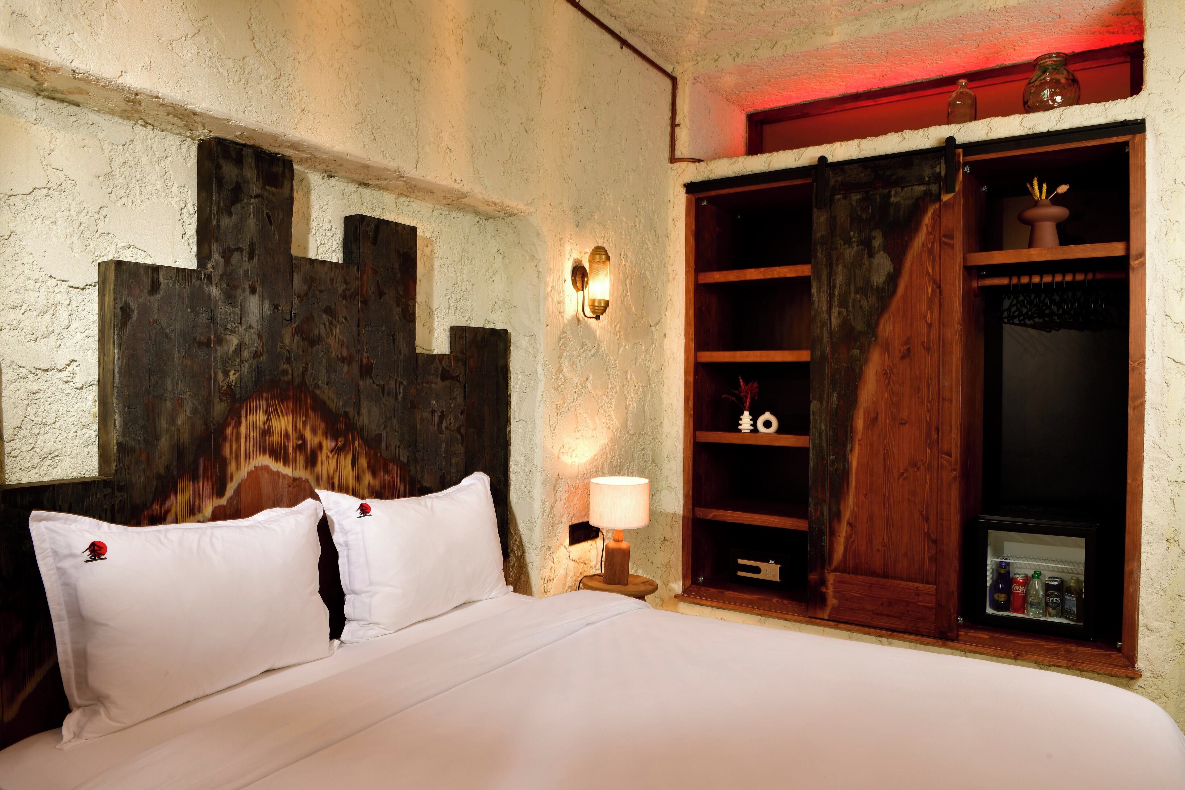 Quarto com cama, mesa de cabeceira, luminária na parede, guarda-roupa de madeira e prateleira com garrafas.