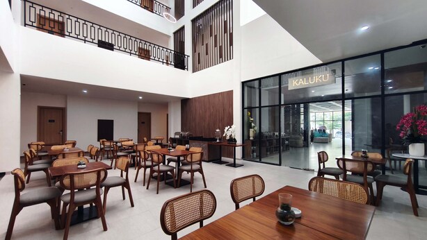 Amplo espaço interno com várias mesas e cadeiras de madeira, restaurante visível chamado "Kaluku" e uma varanda com guarda-corpo.