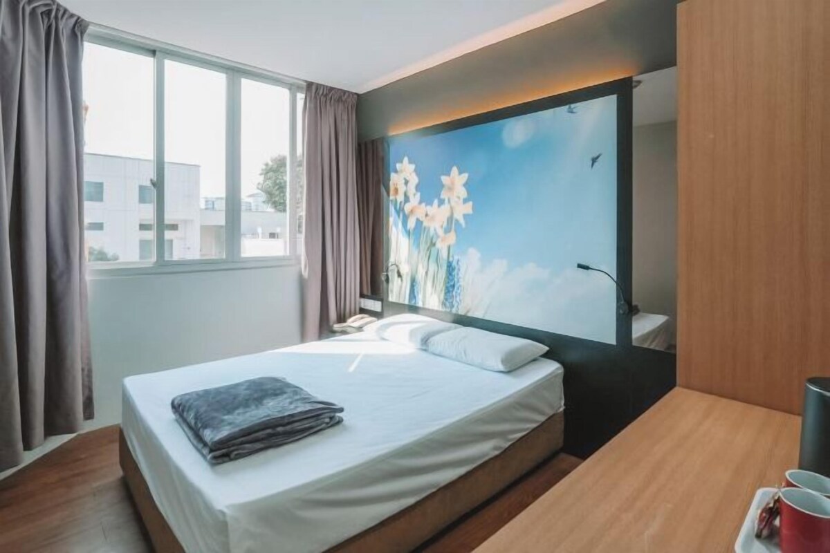Quarto de hotel com cama, mesa de cabeceira com uma chávena, janela com cortinas e um mural na parede.