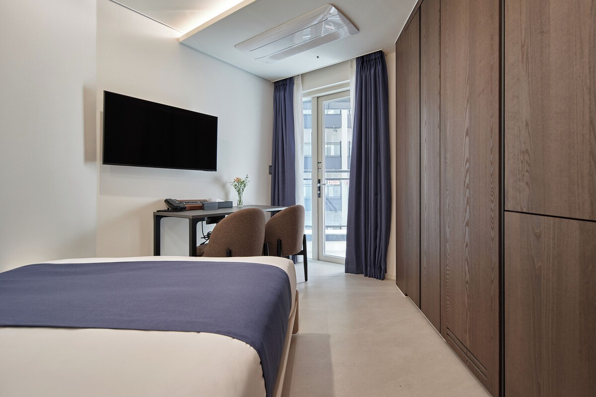Quarto de hotel moderno com cama, escrivaninha e cadeira, televisão de tela plana e um guarda-roupa grande.