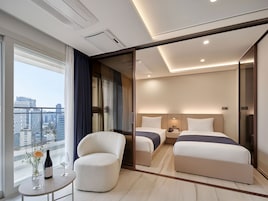 Quarto de hotel moderno com uma cama grande, um espaço de estar com uma poltrona, uma mesa pequena com uma garrafa de vinho e um copo, e vista da cidade por grandes janelas.