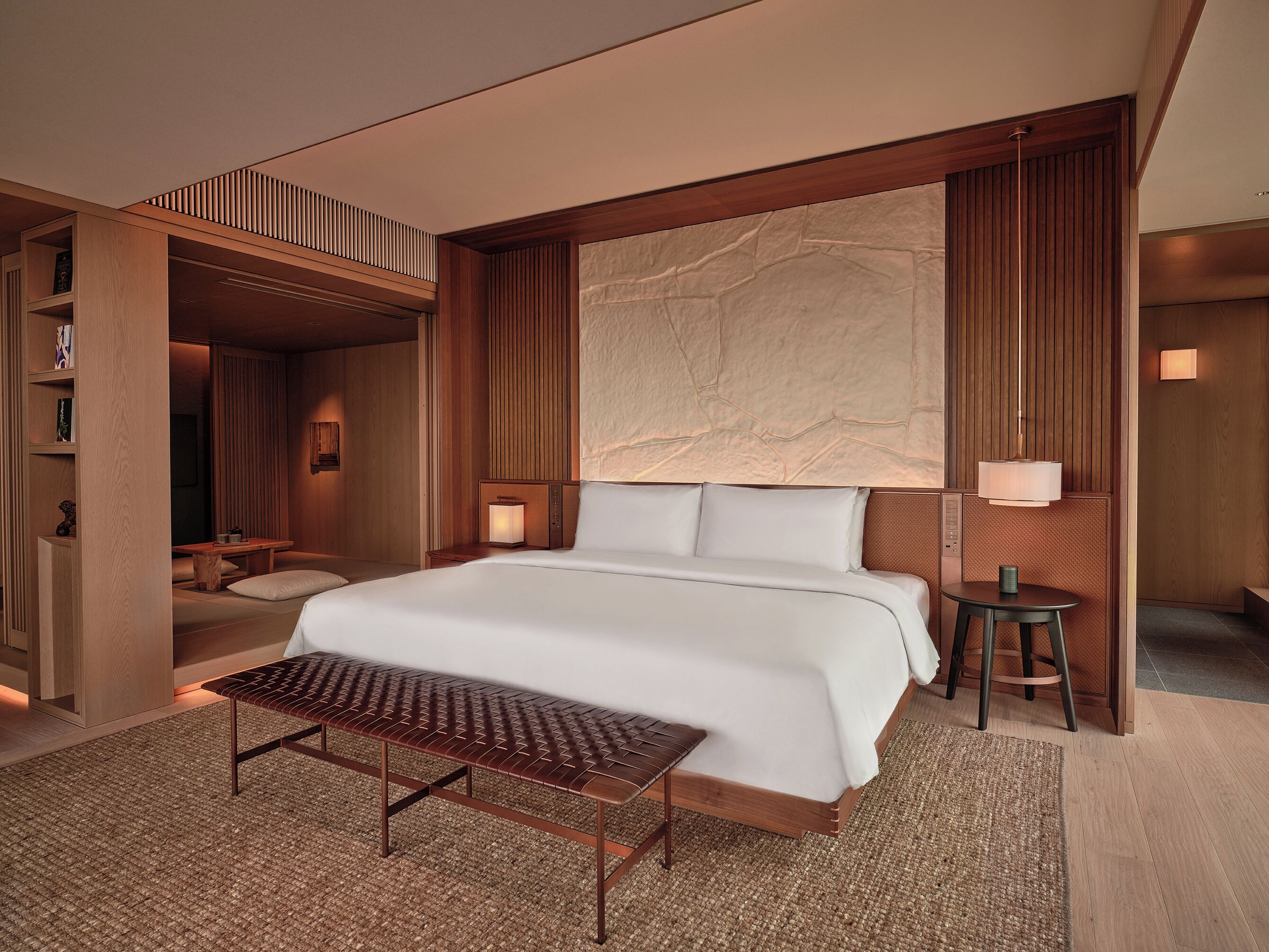 Quarto de hotel moderno com uma cama grande, um banco de madeira, uma mesinha e uma luminária fixada na parede.