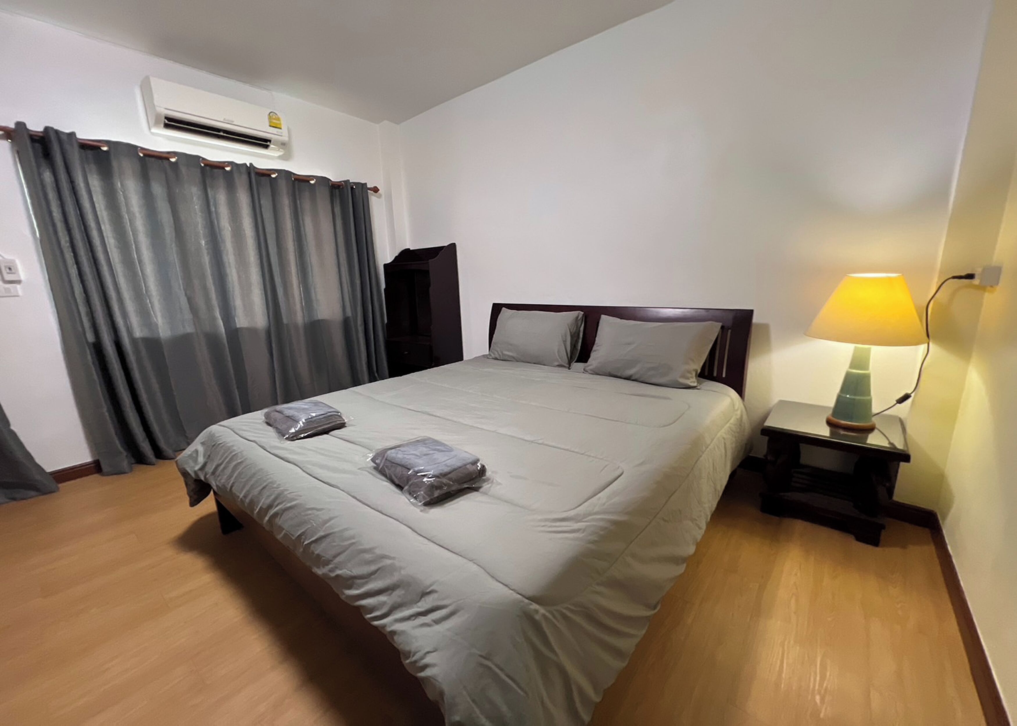 Uma cama bem arrumada com colcha cinza e travesseiros, uma mesa de cabeceira com abajur, e um guarda-roupa.
