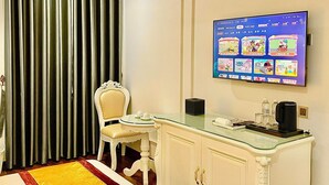 Quarto de hotel com televisão na parede exibindo diversos canais, secretária com tampo de vidro e cafeteira, uma cadeira e uma janela grande com cortinas.
