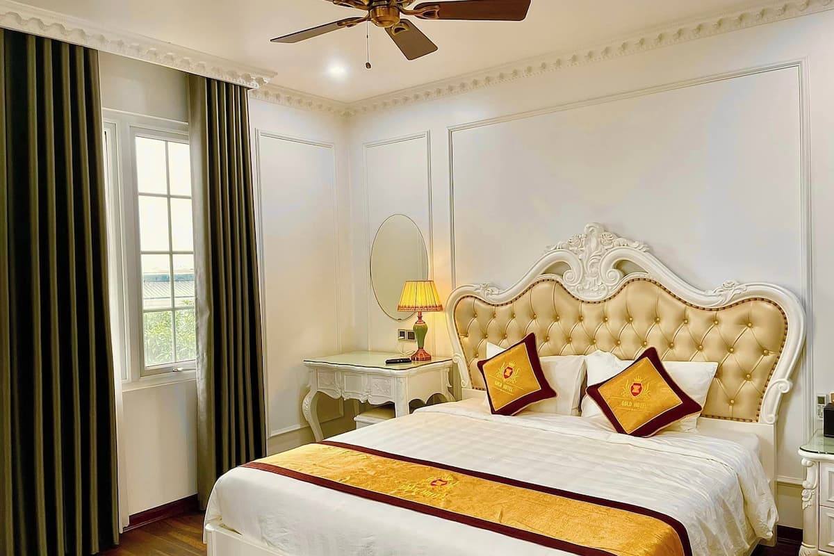 Quarto de hotel com uma cama grande, um ventilador de teto, mesa de cabeceira com luminária e janela com cortinas.