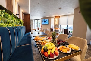 Um saguão de hotel com um sofá azul comprido, uma mesa de jantar com frutas e doces, e uma tela de televisão.