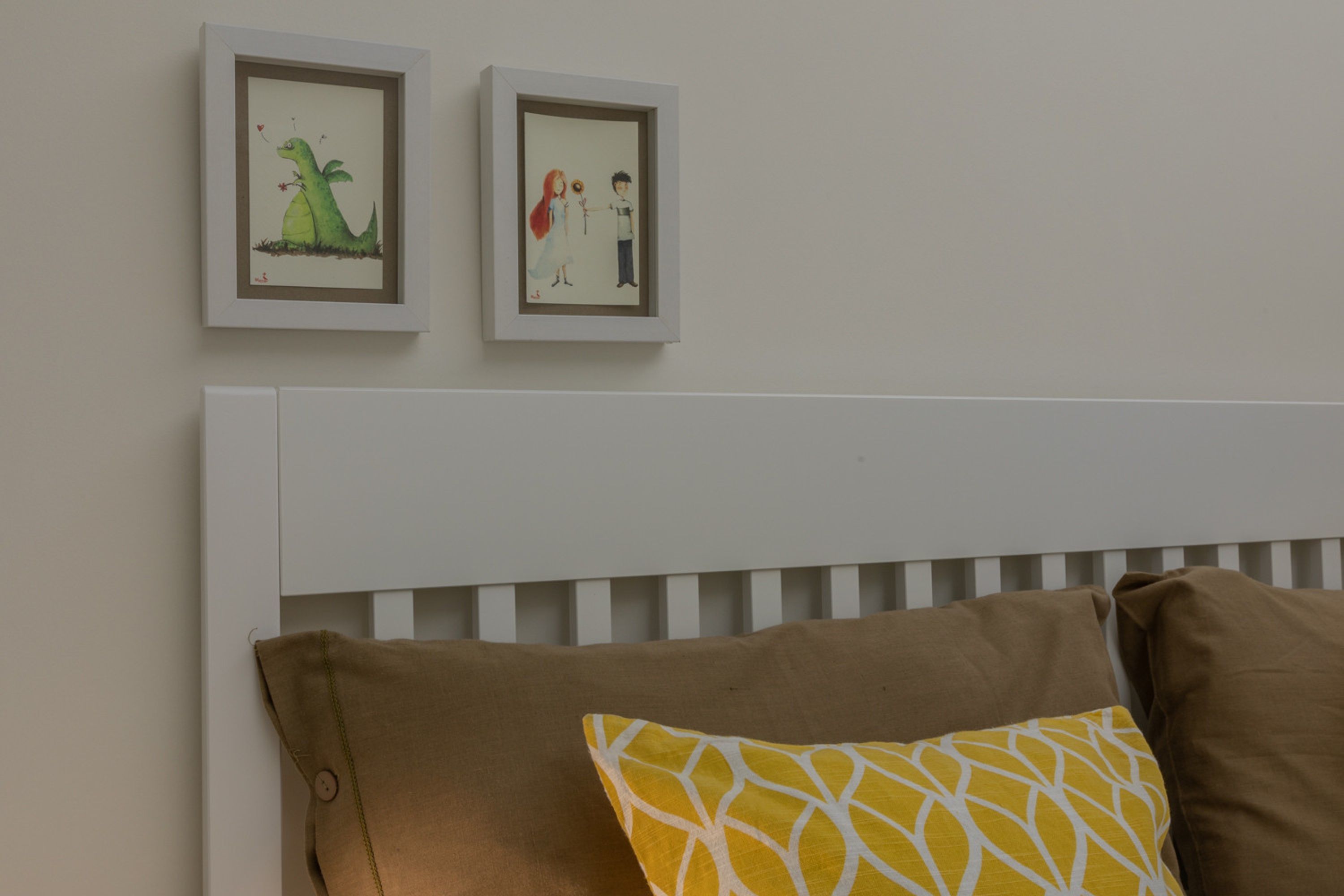 Quarto com cabeceira branca, duas imagens emolduradas na parede e travesseiro marrom e amarelo na cama.
