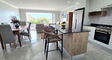 Uma cozinha moderna com ilha central, banquetas, uma mesa de jantar com cadeiras, frigorífico, forno e vista para o exterior.