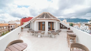 Um terraço no telhado com várias mesas e cadeiras, um pequeno edifício com telhado de azulejos e vista para um horizonte urbano com montanhas ao fundo.