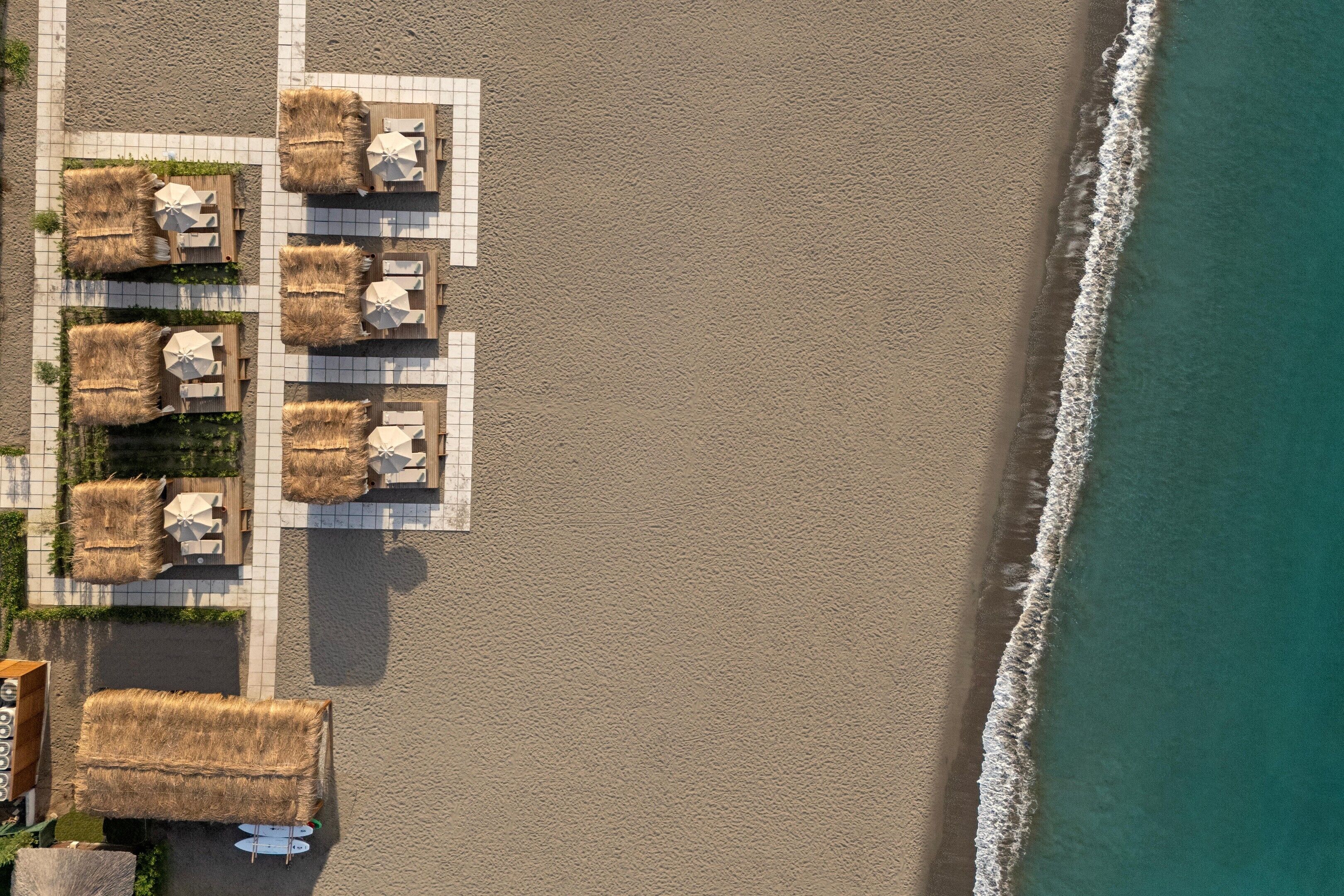 Vista aérea de um hotel à beira-mar com guarda-sóis e mesas de palha, uma praia de areia e o oceano.