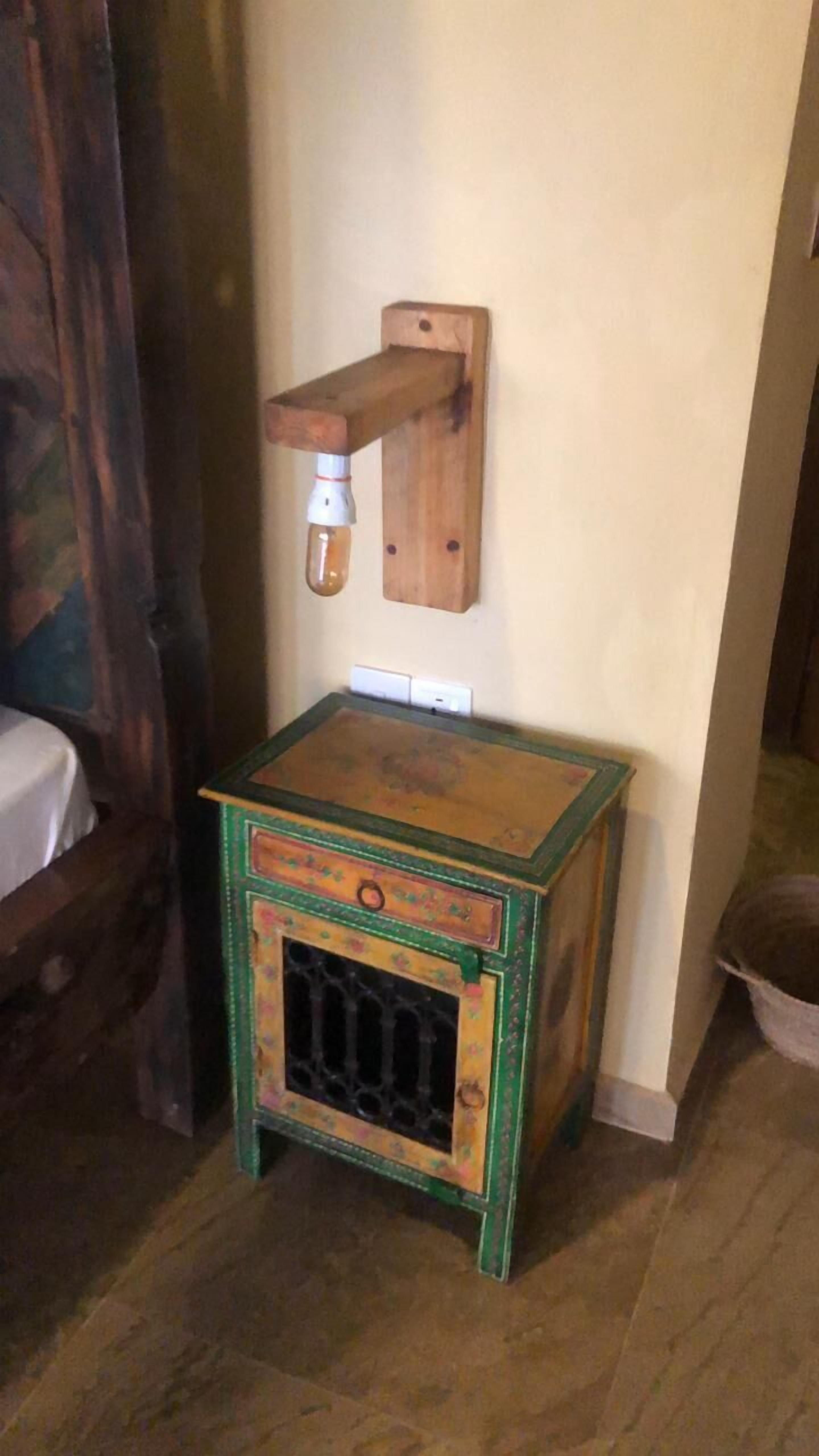 Uma pequena mesa de cabeceira de madeira com uma gaveta, uma prateleira fixada na parede e um aparelho de luz.
