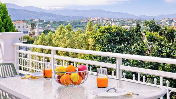 Varanda com mesa posta para o café da manhã, incluindo uma cesta de frutas e copos de suco de laranja, com vista panorâmica de uma cidade e montanhas.