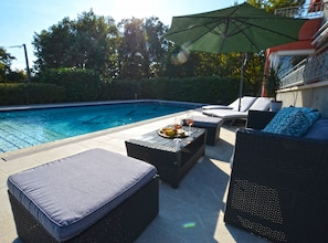Área da piscina com mobiliário de vime, uma mesa com frutas e bebidas, e um guarda-sol.