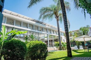 Um hotel moderno com fachada de vidro, área de piscina e vegetação exuberante, incluindo palmeiras e plantas tropicais.