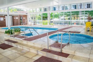 Uma piscina interior com paredes de vidro transparente, escadas de acesso e uma cadeira amarela em primeiro plano.