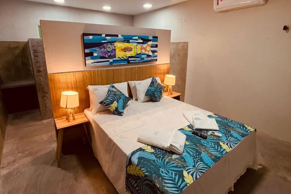 Quarto de hotel com cama, mesa de cabeceira com abajur, ar-condicionado fixado na parede e uma obra de arte colorida sobre a cama.