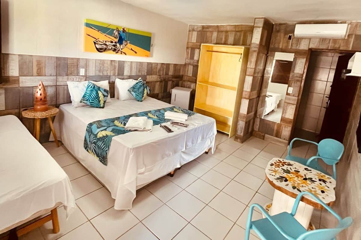 Quarto de hotel com cama, uma pequena mesa com cadeiras, ar condicionado na parede e um quadro sobre a cama.
