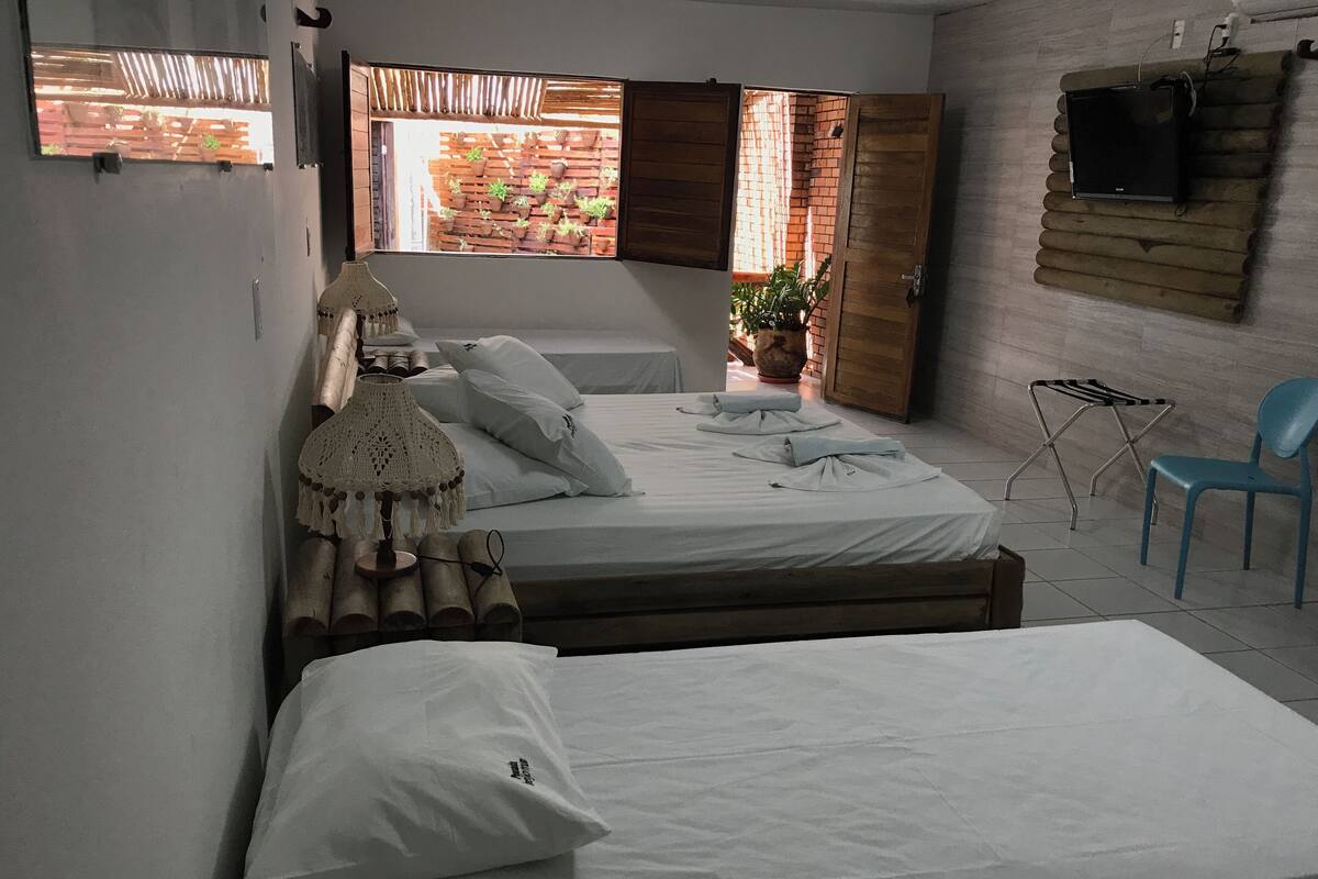 Quarto com duas camas, cabeceira de madeira, televisão fixada na parede e janela com vista para um muro de tijolos e plantas.