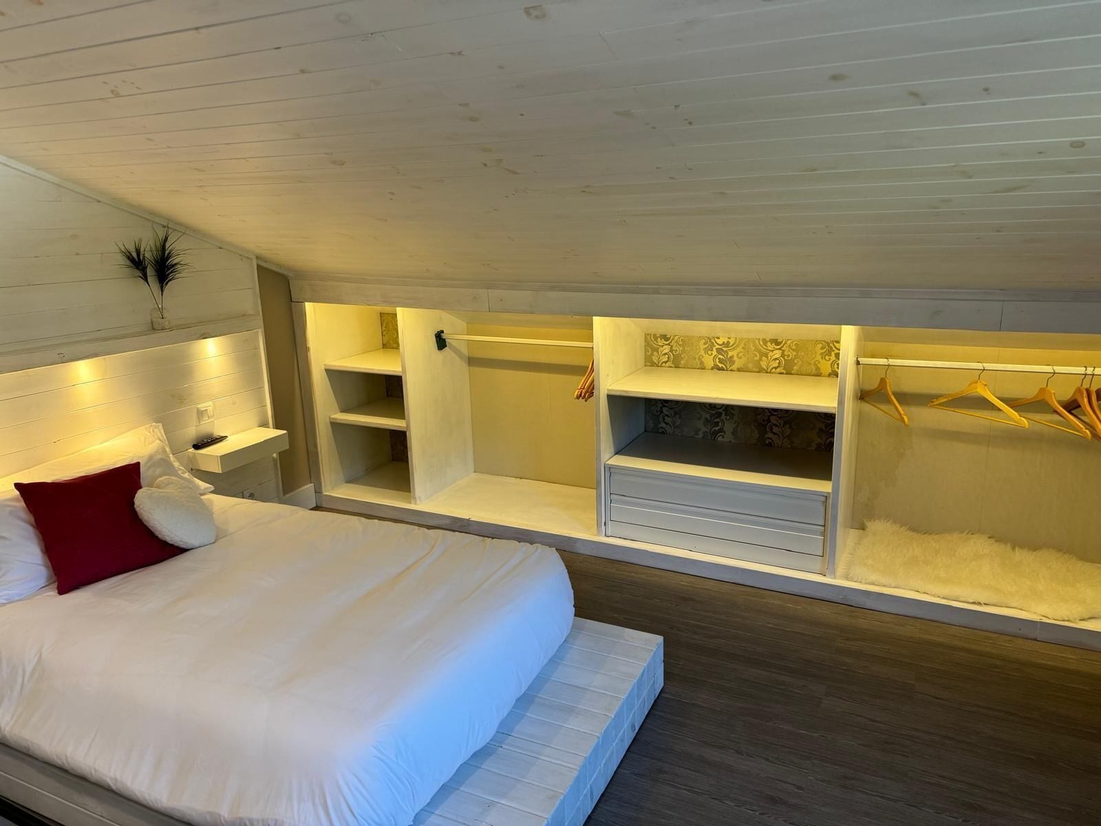 Um quarto com cama, mesa de cabeceira, armário embutido com espaço para pendurar roupas e uma pequena estante.