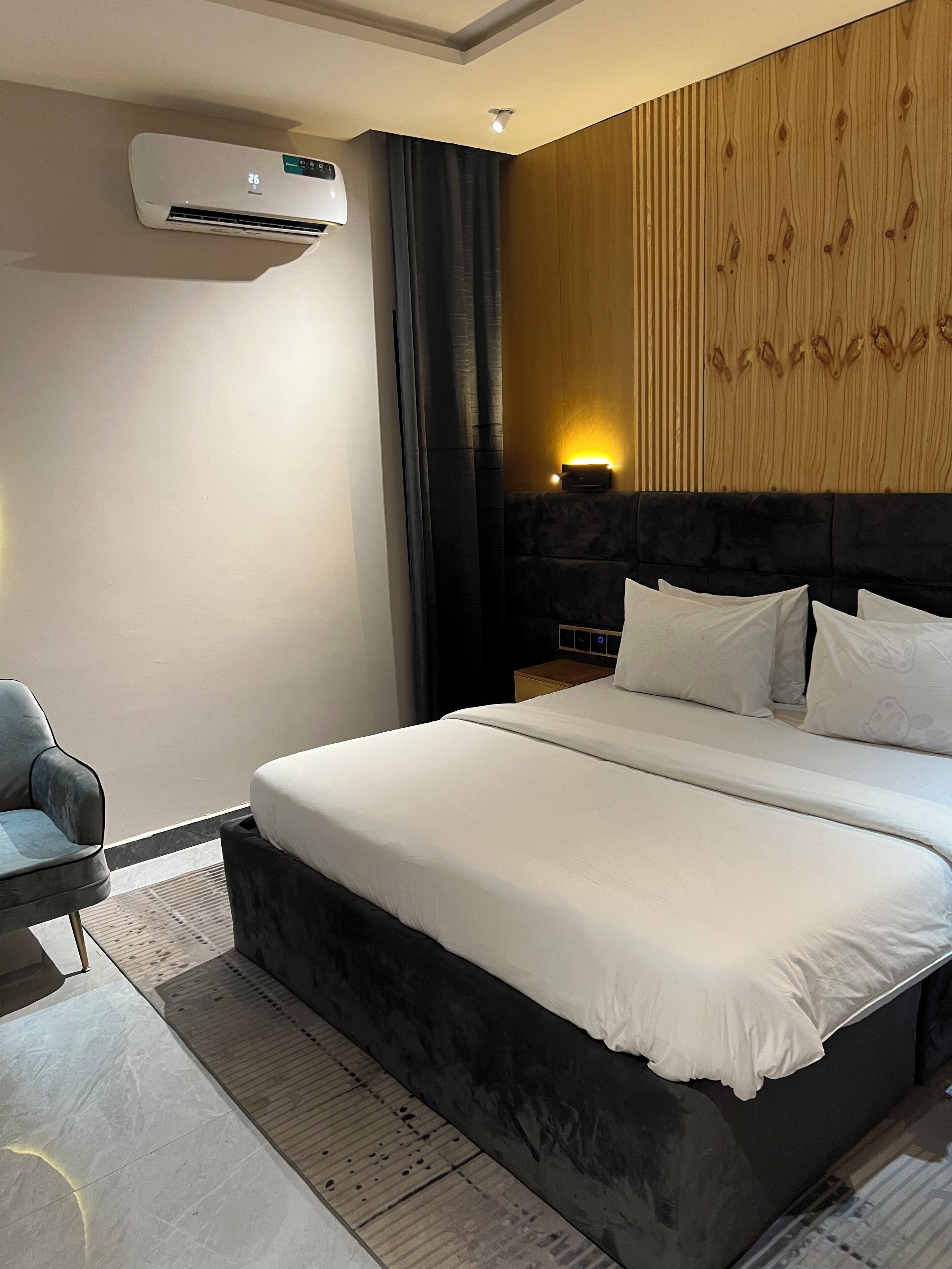 Quarto de hotel moderno com uma cama grande, uma mesinha de cabeceira, uma cadeira e ar condicionado fixado na parede.
