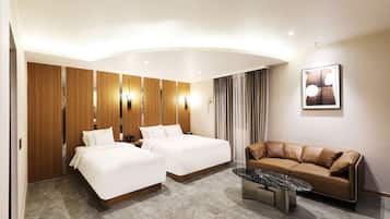 Quarto de hotel com duas camas, um sofá, uma mesa de centro de mármore e uma janela grande com cortinas.