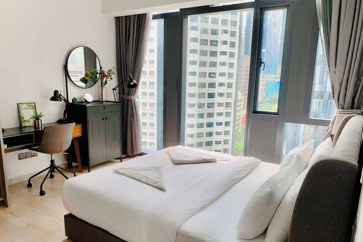 Quarto de hotel moderno com uma cama grande, escrivaninha com cadeira, espelho e vista para edifícios altos.