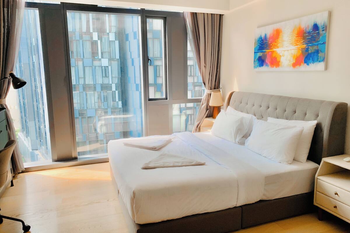 Quarto de hotel com uma cama grande, mesa de cabeceira, abajur e vista da cidade através de janelas do chão ao teto.