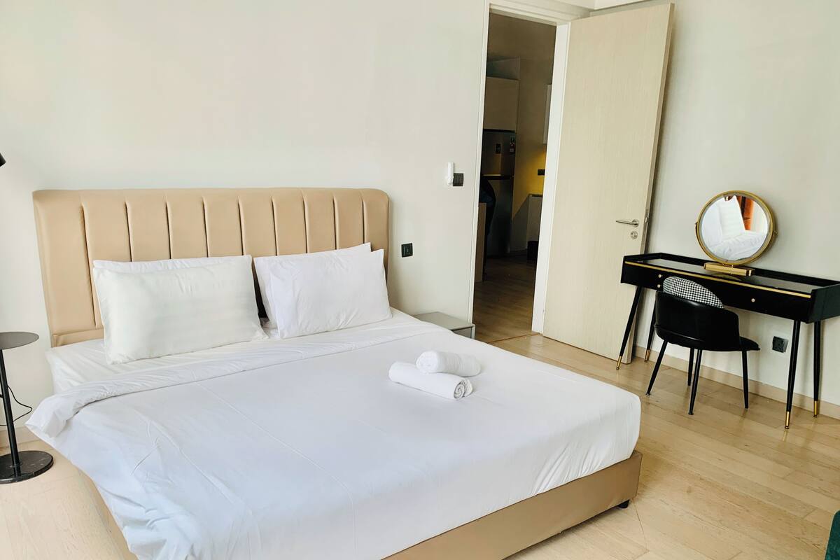 Uma cama bem arrumada com roupa de cama branca e uma toalha dobrada, uma escrivaninha preta com uma cadeira e um espelho na parede.
